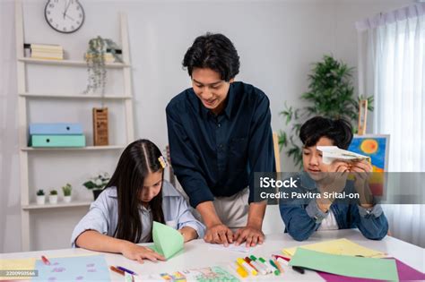 초등학교 미술 수업에서 학생들을 가르치는 아시아 교사 매력적인 남자 교사가 테이블에 앉아 색색의 종이접기 종이를 만드는 귀여운 어린 아이들에게 수업을 설명합니다 10 11