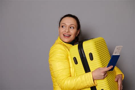 노란 재킷을 입은 쾌활한 여자가 여행 가방을 들고 고립된 회색 배경의 복사 공간을 옆으로 바라보고 있다 여행하다 공휴일에 대한 스톡 사진 및 기타 이미지 Istock