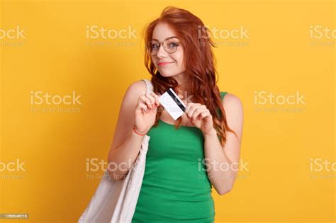 쇼핑백과 신용카드카드를 들고 카메라를 향해 직접 미소짓고 노란 벽에 빨간 머리를 한 여성 쇼핑몰에서 쇼핑을 하고 있는 백인 여성 20 24세에 대한 스톡 사진 및 기타