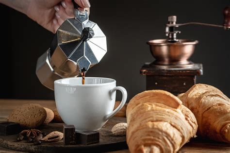 간헐천 커피 메이커의 검은 향기로운 커피를 하얀 컵에 손을 붓고 있습니다 나무 테이블의 머그잔 옆에신선한 크루아상과 커피 그라인더가 있습니다 30 34세에 대한 스톡 사진