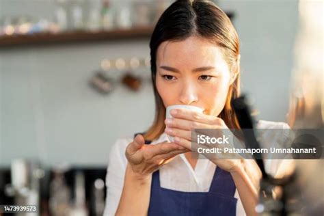 자신감 넘치는 아시아 일본 여성 또는 바리스타가 앞치마를 입고 커피숍 카운터 바에 서서 햇빛 아래 미소로 무장하고 흰색 컵에서 블랙 커피 냄새를 맡고 있습니다 뜨거운 음료의