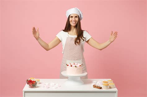 주부 여성 요리사 요리 과자 또는 베이커 앞치마 흰색 T 셔츠 모자 요리사 모자 요리 케이크 또는 테이블에서 컵 케이크 핑크 파스텔 배경에 고립 된 스튜디오에서 복사 공간