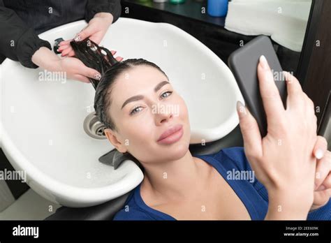 Brunette Woman Looks At Smartphone While Hairdresser Is Busy Washing Her Hair At Beauty Salon