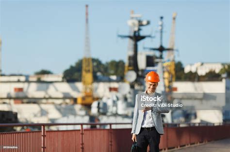 잘생긴 젊은 이루어지지 성공적인 사업가 회색 양복과 보호 건설 오렌지 헬멧 지주 케이스 시계 시간을 찾고 화물선에 대 한 항구에 산책 및 배경 크레인 20 29세에 대한