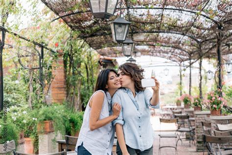 Lesbian Couple In Plant Covered Archway Using Smartphone To Take Selfie