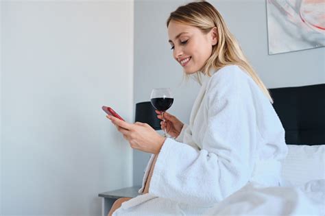 현대 아늑한 호텔에서 휴대 전화를 사용하여 행복하게 레드 와인 한 잔을 들고 흰색 목욕 가운에 젊은 예쁜 미소 금발 여자 20 24세에 대한 스톡 사진 및 기타 이미지