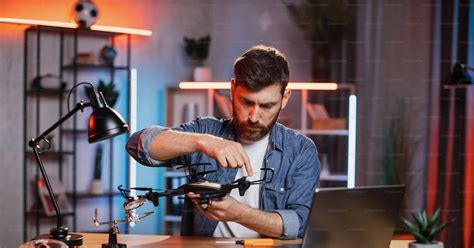 집에서 현대 쿼드콥터를 수리하기 위해 다양한 도구를 사용하는 집중된 수염 난 남자 데님 셔츠를 입은 젊은 백인 남자가 혼자 비행 드론을 고치고 있다 사진