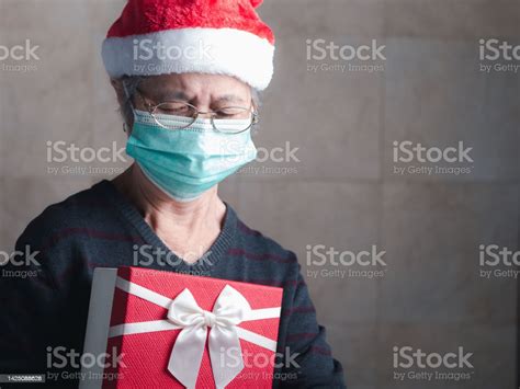 코로나 바이러스로 인해 외과 용 마스크를 착용하고 산타 클로스 모자를 쓰고 포옹을하고 빨간 선물 상자를보고있는 노인 여성 12월에 대한 스톡 사진 및 기타 이미지 Istock