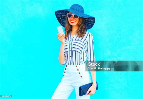 행복 한 여자는 밀 짚 모자를 쓰고 스마트폰 사용 하 고 화이트 바지와 핸드백 클러치 화려한 파란색 배경 위에 가방에 대한 스톡 사진 및 기타 이미지 Istock