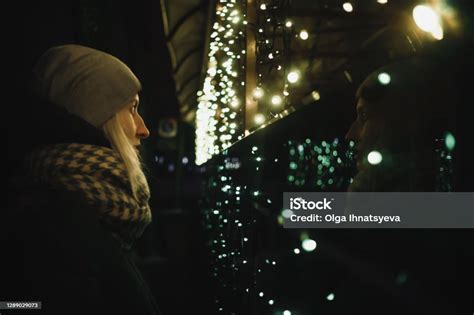 모자를 쓰고 아래 재킷과 볼륨감 있는 스카프를 입은 젊은 금발 여성이 장식된 상점 창문의 화환 카페 클로즈업 창에서 어둠 속에서