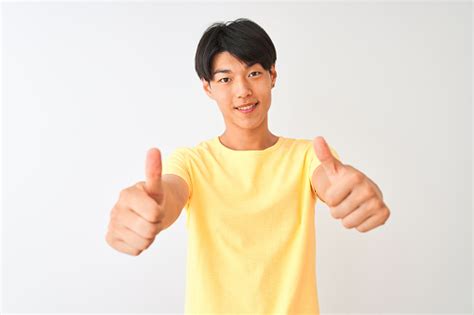 노란색 캐주얼 티셔츠를 입은 중국 남자는 손으로 긍정적 인 제스처를 하고 엄지 손가락으로 미소를 지으며 성공을 위해 행복한 흰색 배경 위에 서 있습니다 우승자 제스처 T