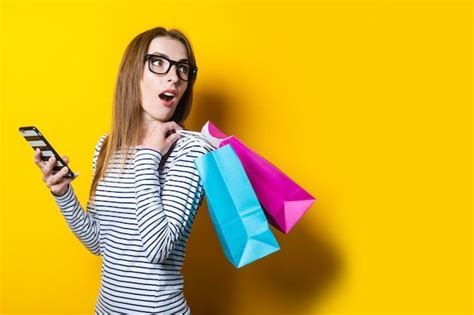 Удивленная молодая женщина оглядывающаяся назад держа телефон и хозяйственные сумки на желтом