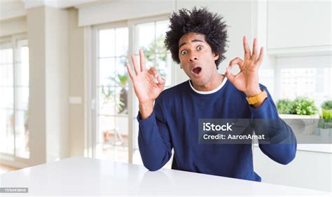 캐주얼 한 스웨터를 입고 집에 앉아있는 젊은 아프리카 계 미국인 남자는 손가락으로 확인 승인 기호를하고 놀랍고 충격을 받았습니다