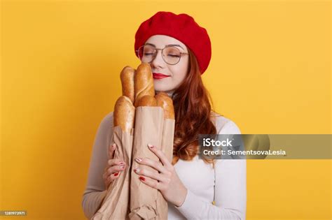 노란 배경에 빵종이 봉투를 들고 있는 아름다운 빨간 머리의 젊은 여성은 만족스러운 소녀는 눈을 감고 캐주얼 셔츠와 베레모를 입은 아가씨와 신선한 향기로운 긴 덩어리 냄새가