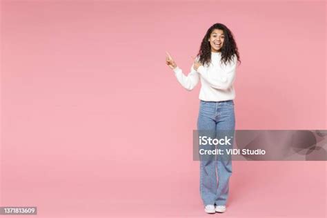 파스텔 핑크 배경 스튜디오 초상화에 고립 된 복사 공간 공간에 흰색 니트 스웨터 청바지 포인트 인덱스 손가락 옆으로 전체 길이 젊은 행복 미소 아프리카 계 미국인 여성 20