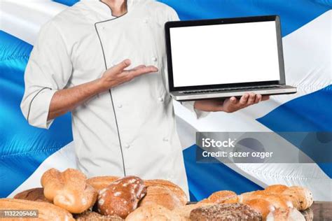 스코틀랜드 국기와 빵 배경에 노트북을 들고 스코틀랜드 베이커 요리사가 복사 공간에 대 한 균일 한 포인팅 빈 화면을 착용 고급 요리에 대한 스톡 사진 및 기타 이미지