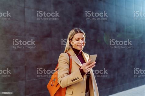 매력적인 백인 미소 금발 여자 코트와 지갑 야외서 동안 스마트 폰을 들고 배경은 어두운 벽입니다 가방에 대한 스톡 사진 및 기타 이미지 Istock