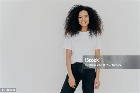 곱슬 곱슬 한 어두운 머리를 가진 미소 짓는 여자의 사진 슬림 한 그림을 가지고 흰색 티셔츠와 검은 청바지를 입고 기쁜 얼굴 표정 흰색 배경에 대한 모델 광고 콘텐츠에 대한