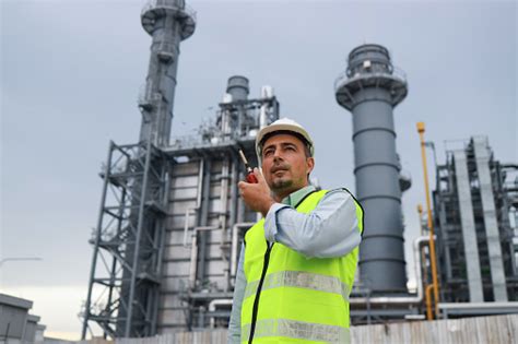 밝은 녹색 반사 조끼와 흰색 안전모를 착용하고 화력 발전소 현장에서 무전기를 사용하여 작업하는 똑똑한 선임 남성 엔지니어의 초상화 기술자에 대한 스톡 사진 및 기타 이미지