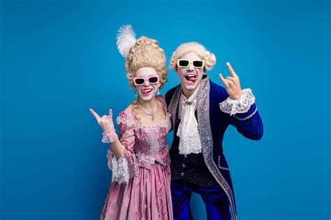 바로크 양식의 의상과 선글라스를 낀 장난기 넘치는 커플이 강렬한 파란색 배경에서 역사적인 우아함과 현대가 조화를 이루며 포즈를 취하고 있습니다 16 17 살에 대한 스톡