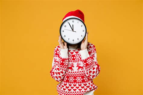 빨간 스웨터 크리스마스 모자를 쓴 젊은 산타 여성 20 대는 노란색 배경 스튜디오 초상화에 고립 된 시계와 얼굴을 덮고 손에 들고 있습니다 새해 축하 메리 홀리데이 컨셉