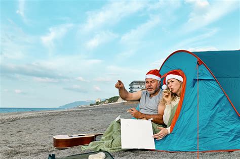 산타 모자를 쓴 중년 부부가 겨울 해변에서 크리스마스 피크닉을 즐기고 있다 턱수염 난 남자와 예쁜 금발의 여자가 텐트에 앉아 피자를 먹고 이야기한다 12월에 대한 스톡 사진