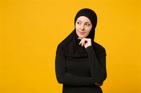 웃 고 아름 다운 젊은 아라비아 무슬림 여자 히 잡 블랙 옷 노란 벽 배경 스튜디오 초상화에 고립 된 카메라를 옆으로 찾고 사람들 종교적 라이프 스타일 개념입니다 복사
