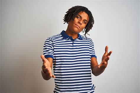 스트라이프 블루 폴로를 입고 고립된 흰색 배경에 서있는 공포를 가진 아프로 남자는 단서가 없고 팔을 벌려 혼란스러워하며 아무