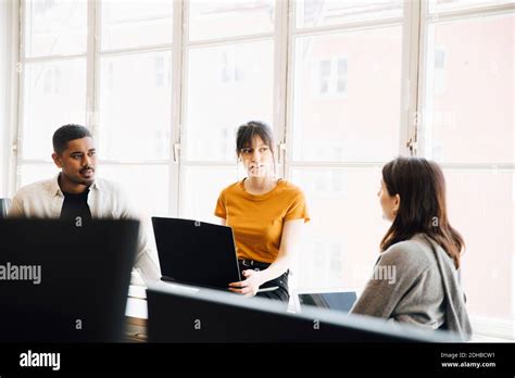 Female Software Engineer Explaining To Coworkers While Sitting Against Window In Office Stock