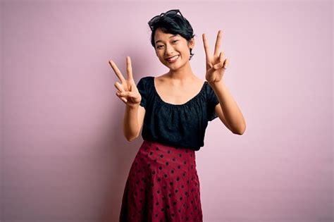 고립 된 분홍색 배경 위에 서있는 캐주얼 한 드레스를 입은 젊은 아름다운 아시아 소녀는 승리의 표지판을하고있는 양손의 손가락을 보여주는 혀로 미소 짓고 있습니다 2번