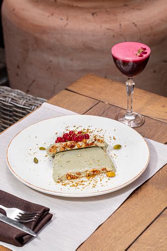 라즈베리 거품과 장미 꽃 봉오리를 곁들인 버몬트 줄기가 높은 유리에 담긴 음료 나무 탁자 위에 서 있다 근처에는 피스타치오 아이스크림과 붉은 건포도 접시가 있습니다 0명에