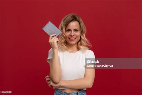 빨간 배경에 서 흰색 티셔츠에 곱슬 금발 머리를 가진 여자의 초상화입니다 모델은 카메라를 미소 짓고 파란색 여권을 들고 있습니다 T 셔츠에 대한 스톡 사진 및 기타 이미지
