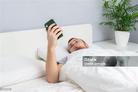 수염을 기른 남자는 집 침대에서 전화를 사용합니다 침대에서 일어나지 않고 물건을 쇼핑하십시오 가제트에 대한 사람들의 의존 개념 인터넷 중독 전화에 대한 의존성 가제트 중독