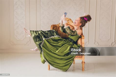 우아한 역사적인 의상을 입은 젊은 여성이 고급스러운 바로크 양식의 인테리어 환경에서 과거와 현재의 현대적인 비교를 즐기고 있습니다 20 24세에 대한 스톡 사진 및 기타