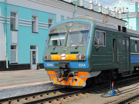 a green and orange train traveling down tracks next to a blue building ...