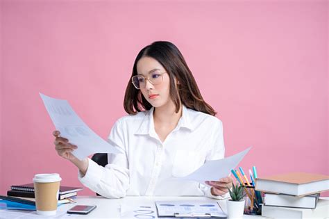 안경을 쓰고 흰 셔츠를 입은 젊은 아시아 여성이 흰색 책상에 앉아 있다 그녀는 사무실에서 노트북으로 문서를 들고 있다 N 파스텔 핑크 배경에 격리 30 34세에 대한 스톡
