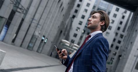 자신감있는 사업가 정장을 입은 자신감 넘치는 청년이 스마트폰을 사용하고 도시 풍경을 배경으로 야외에 서 있는 동안 시선을 돌리고 있다 사진 Unsplash의 양복 이미지