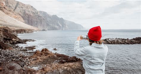 빨간 모자를 쓴 젊은 여성이 바위가 많은 바다 해안에서 여행을 즐기며 전화로 숨막히는 풍경을 촬영하고 있습니다 스페인 테네리페 섬 여행 사진 Unsplash의 청녀 이미지