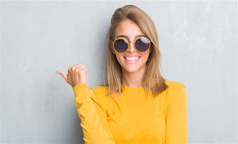 아름 다운 젊은 여자를 가리키는 및 행복 한 얼굴은 웃 고 옆까지 엄지 보여주는 복고풍 선글라스를 착용 하는 그런 지 회색 벽을 통해 서 가리키기에 대한 스톡 사진 및 기타