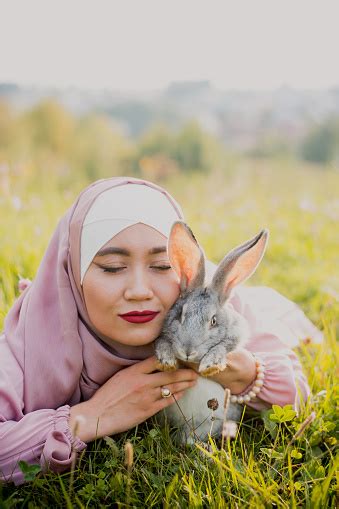 무슬림 여성이 들판에 누워 회색 토끼를 품에 안았다 분홍색 드레스와 스카프 진주 팔찌를 입은 무슬림 여성의 초상화 부드러움 감정에 대한 스톡 사진 및 기타 이미지 Istock