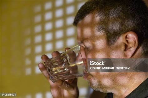한 남자가 탐욕스럽게 유리잔에서 물을 마셨다 벽에 부딪히는 중년 남성의 초상화 벽과 남자의 얼굴에 반복되는 인물의 빛과 그림자 패턴 측면보기 낮은 키 식수에 대한 스톡 사진
