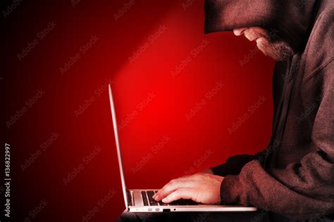 Male Computer Hacker Wearing A Hooded Top Leaning Over A Laptop In The Dark The Screen Light