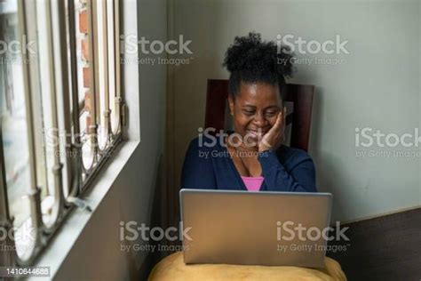 창문 옆에 앉아있는 노트북에서 스트리밍을 통해 자신이 좋아하는 시리즈의 장을 보는 흑인 여성 50 59세에 대한 스톡 사진 및 기타 이미지 Istock