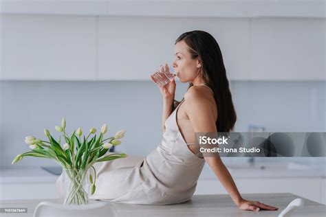 이브닝 드레스를 입은 사려 깊은 스페인 젊은 여성이 튤립으로 가득 찬 꽃병이 물을 마시며 식탁에 앉아 있습니다 긴 머리를 가진 아름다운 백인 여성은 집에서 하루를 시작합니다