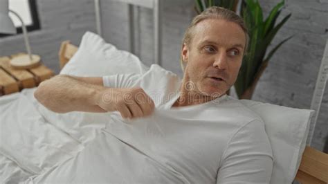 Mature Caucasian Man With Blonde Hair In A White Shirt Sitting On A Bed