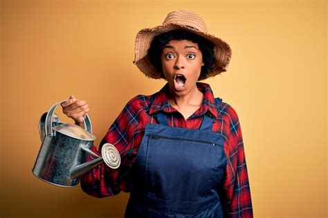 앞치마와 모자를 쓰고 곱슬 머리를 한 젊은 아프리카 계 미국인 농부 여성은 깜짝 얼굴로 충격에 두려워할 수 있으며 두려움과 두려움의 표정으로 흥분할 수 있습니다 감정에 대한