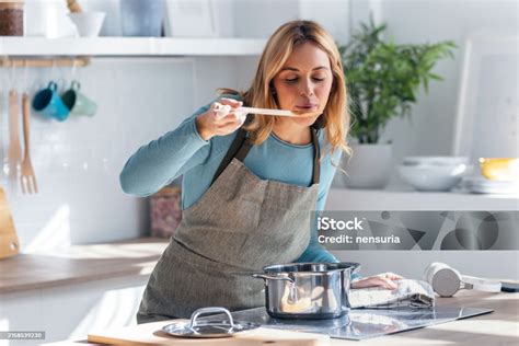 집에서 부엌에서 음식을 맛보기 위해 숟가락을 불면서 캐서롤에서 건강한 음식을 요리하는 아름 다운 여자 25 29세에 대한 스톡 사진 및 기타 이미지 Istock