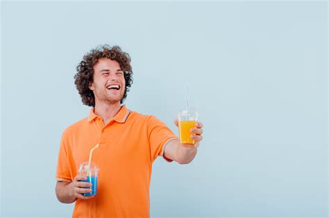 곱슬 머리 미소와 잘 생긴 행복한 남자 오렌지 셔츠 고립 된 파란색 배경 긍정적 인 얼굴 감정 복사 공간을 입고 주스와 두 개의 플라스틱 컵을 보유 곱슬 머리에 대한 스톡