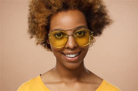 근접 촬영 얼굴만 잘생긴 아프리카계 미국인 여자의 사진에 고립 된 베이지색 배경 노란색 위쪽 및 보석 긍정적으로 미소를 입고 유행 안경을 통해 카메라를 똑바로 보고 T