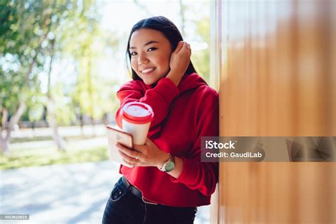 스마트폰과 카페인 음료를 들고 도시의 도시 환경에 서서 카메라를 향해 웃고 있는 예쁜 아시아 밀레니얼 세대의 절반 길이 초상화 브라우징을 위해 디지털 기술을 들고 있는